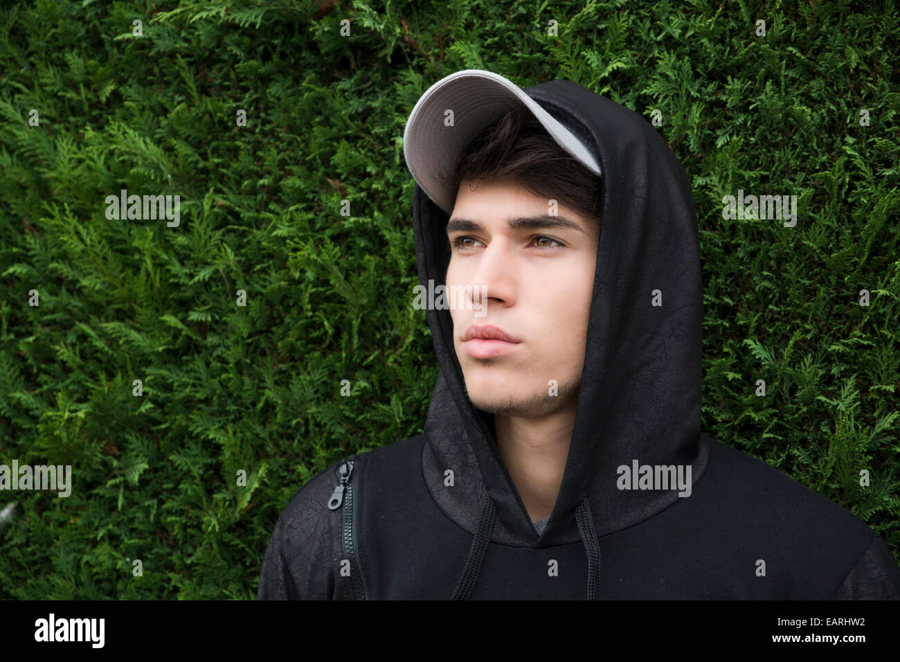 Attractive young man with hoodie and baseball cap against green bush ...