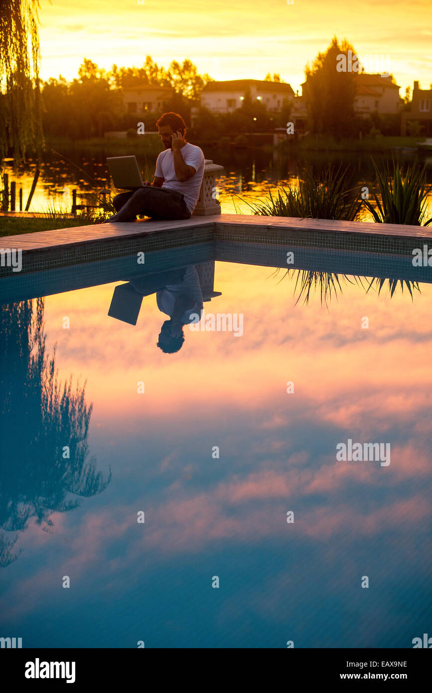 Man using laptop computer next to resort swimming pool as sun sets ...