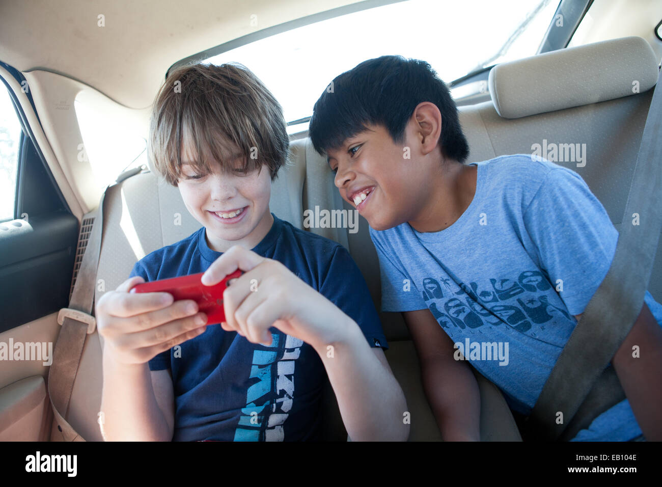 Two ten year old boys watch a video on a phone, back seat of a car ...