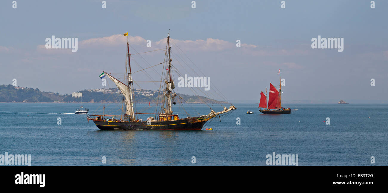 A twin masted sailing ship is at anchor with a red sailed boat in the ...