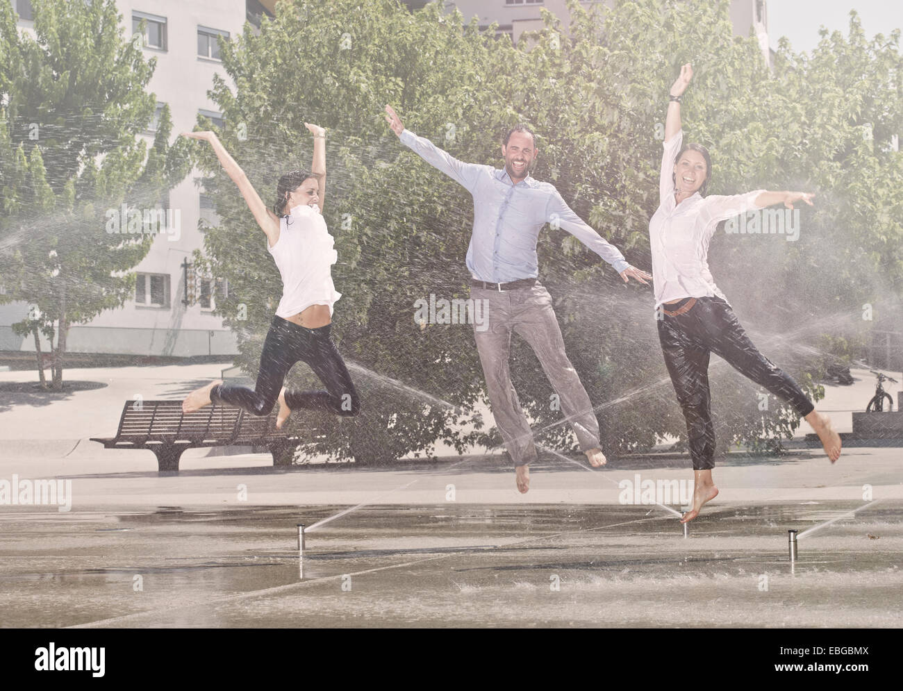 Two women and one man jumping in the park, between water sprinklers ...
