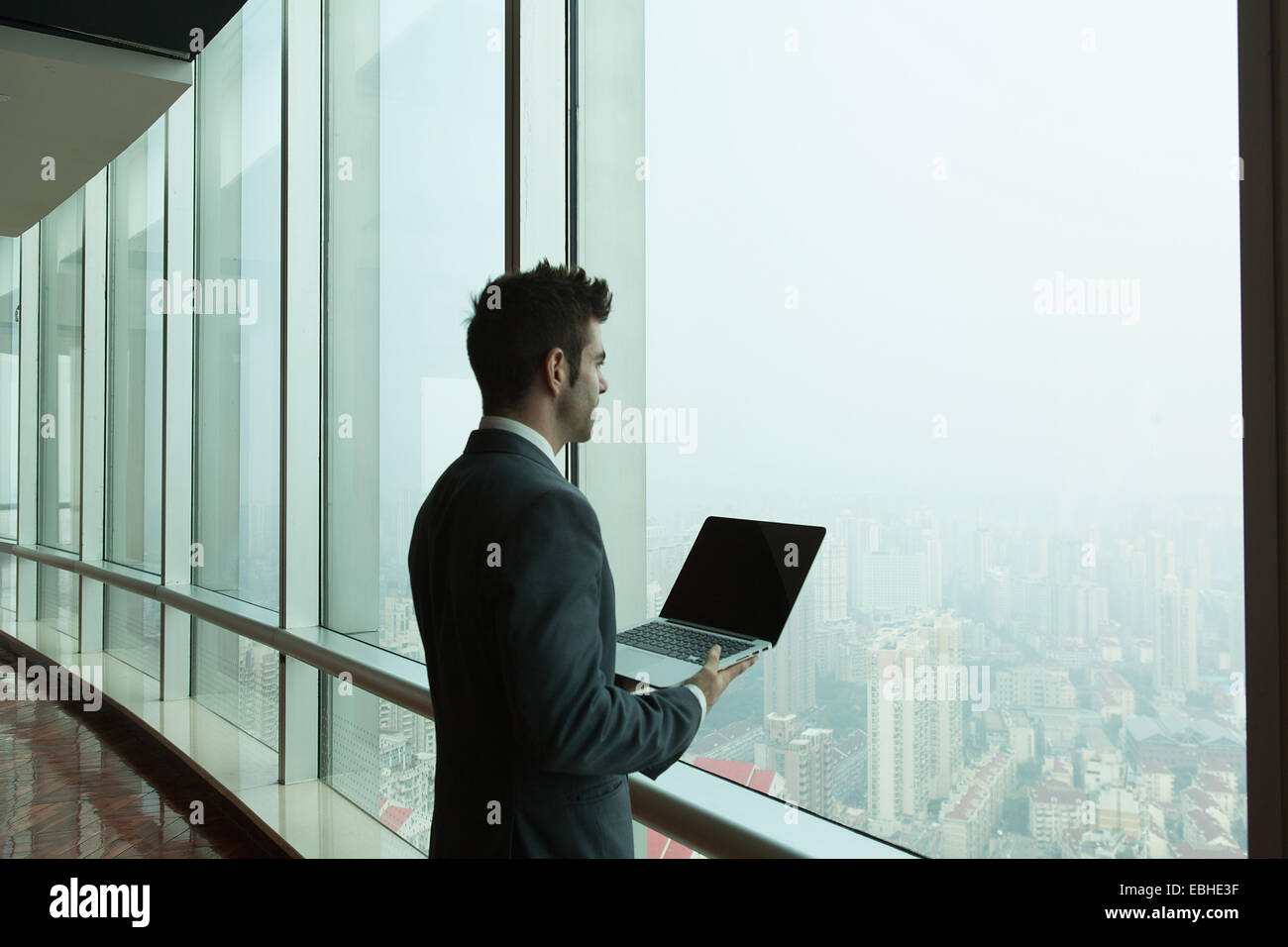 Young businessman with laptop looking out of skyscraper office window ...