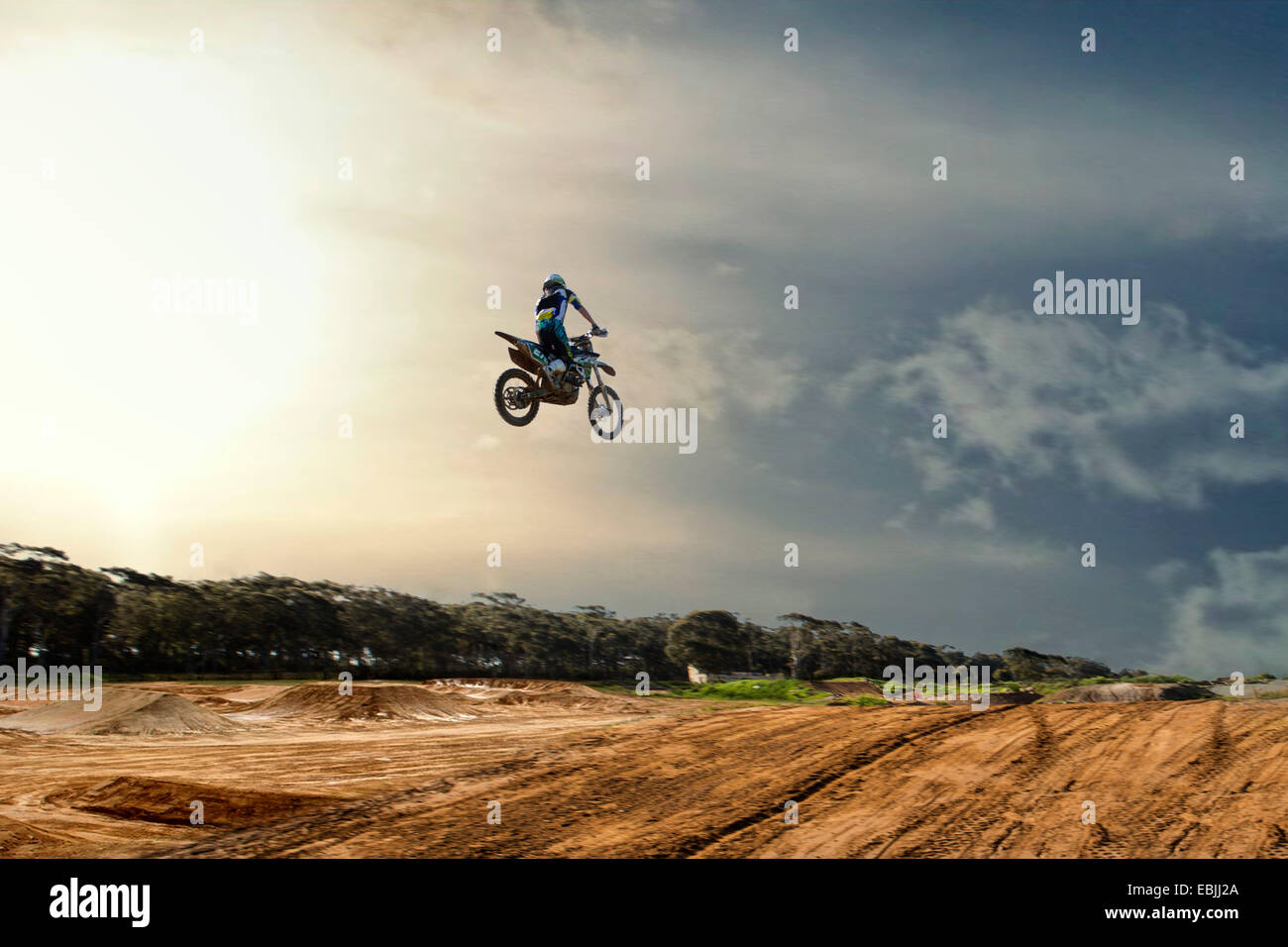 Silhouetted young male motocross racer jumping mid air over mud track ...