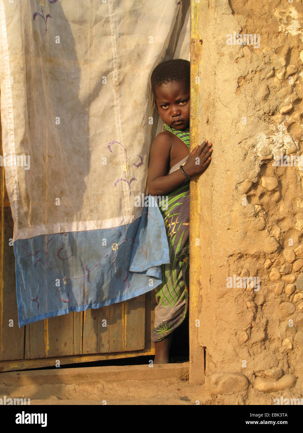 little girl looking out of the wooden entrance door of a simple mud ...