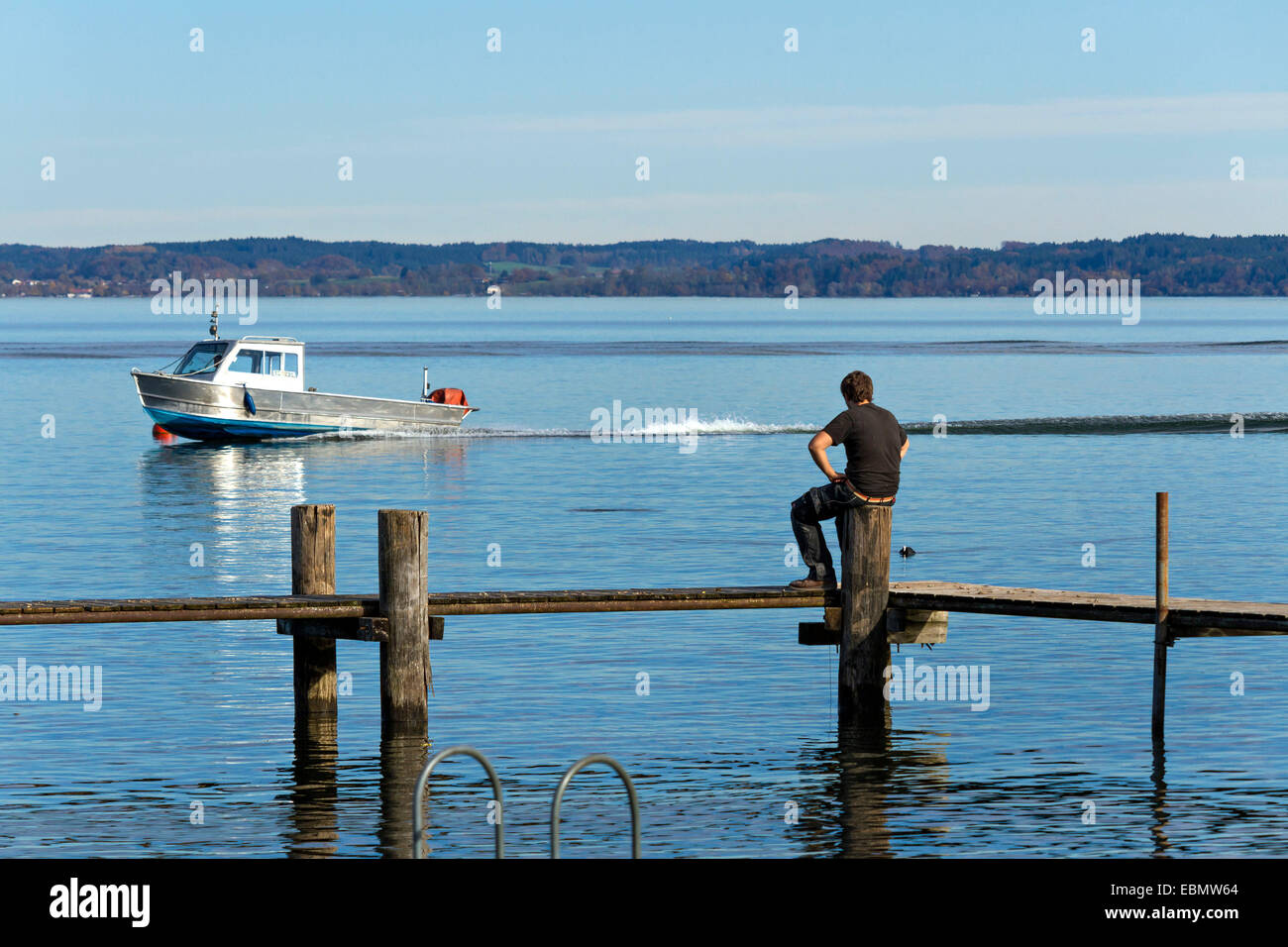 Man sitting on wood pylon as power boat passes, Fraueninsel, Chiemsee, Chiemgau, Upper Bavaria ...