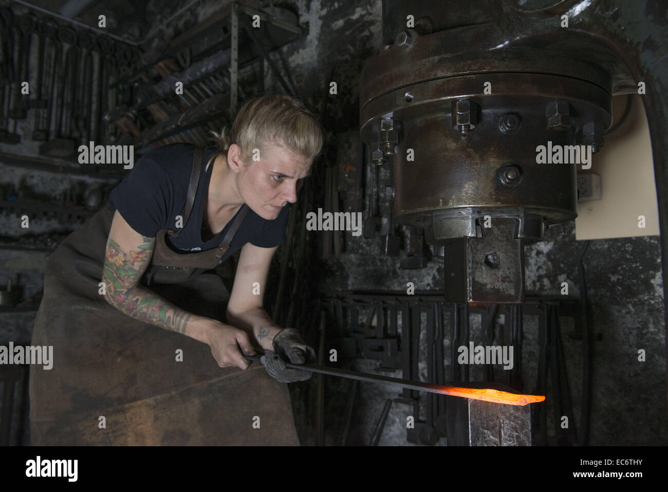 female blacksmith works a glowing piece of steel with steam hammer ...