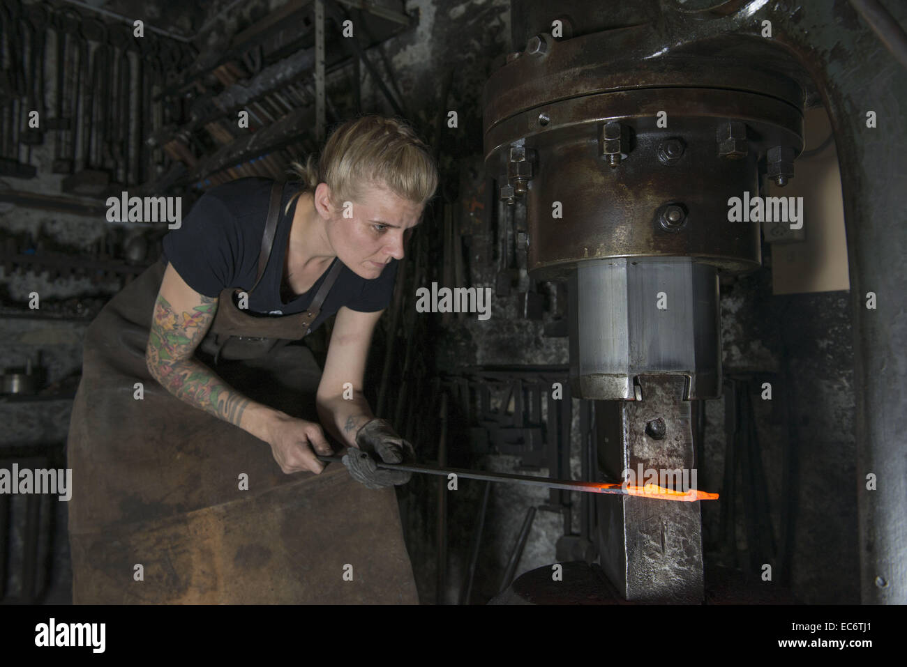 female blacksmith works a glowing piece of steel with steam hammer ...