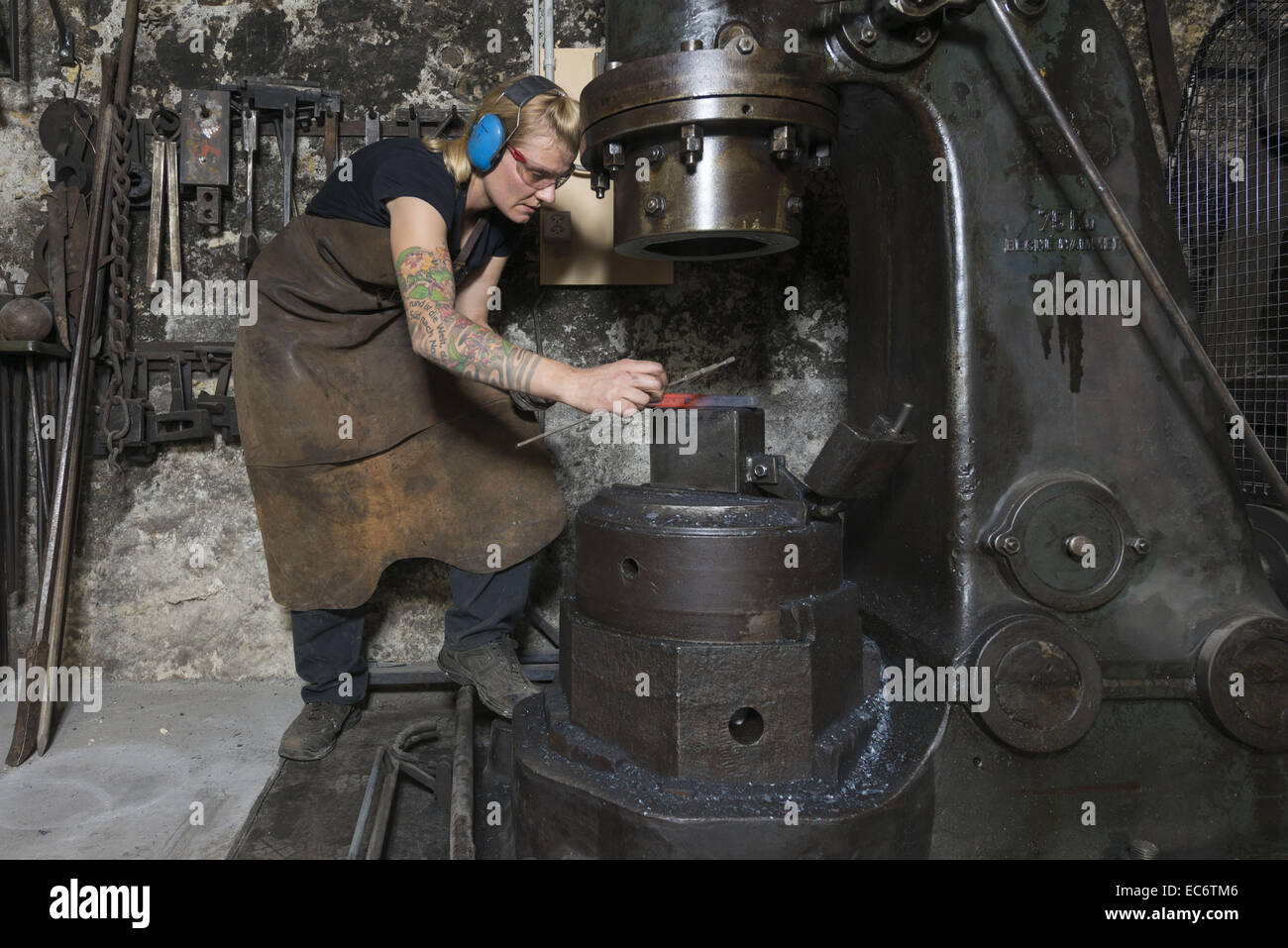 female blacksmith measuring a glowing piece of steel, steam hammer ...