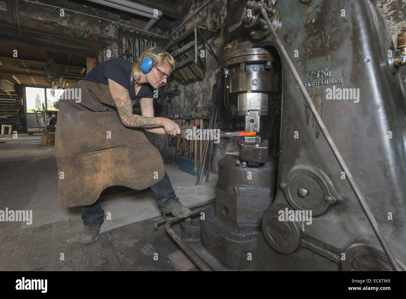 female blacksmith works a glowing piece of steel with steam hammer ...