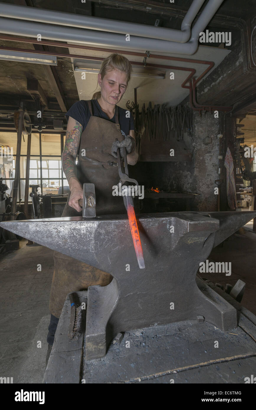 female blacksmith inspecting a redhot piece of steel on anvil, window ...