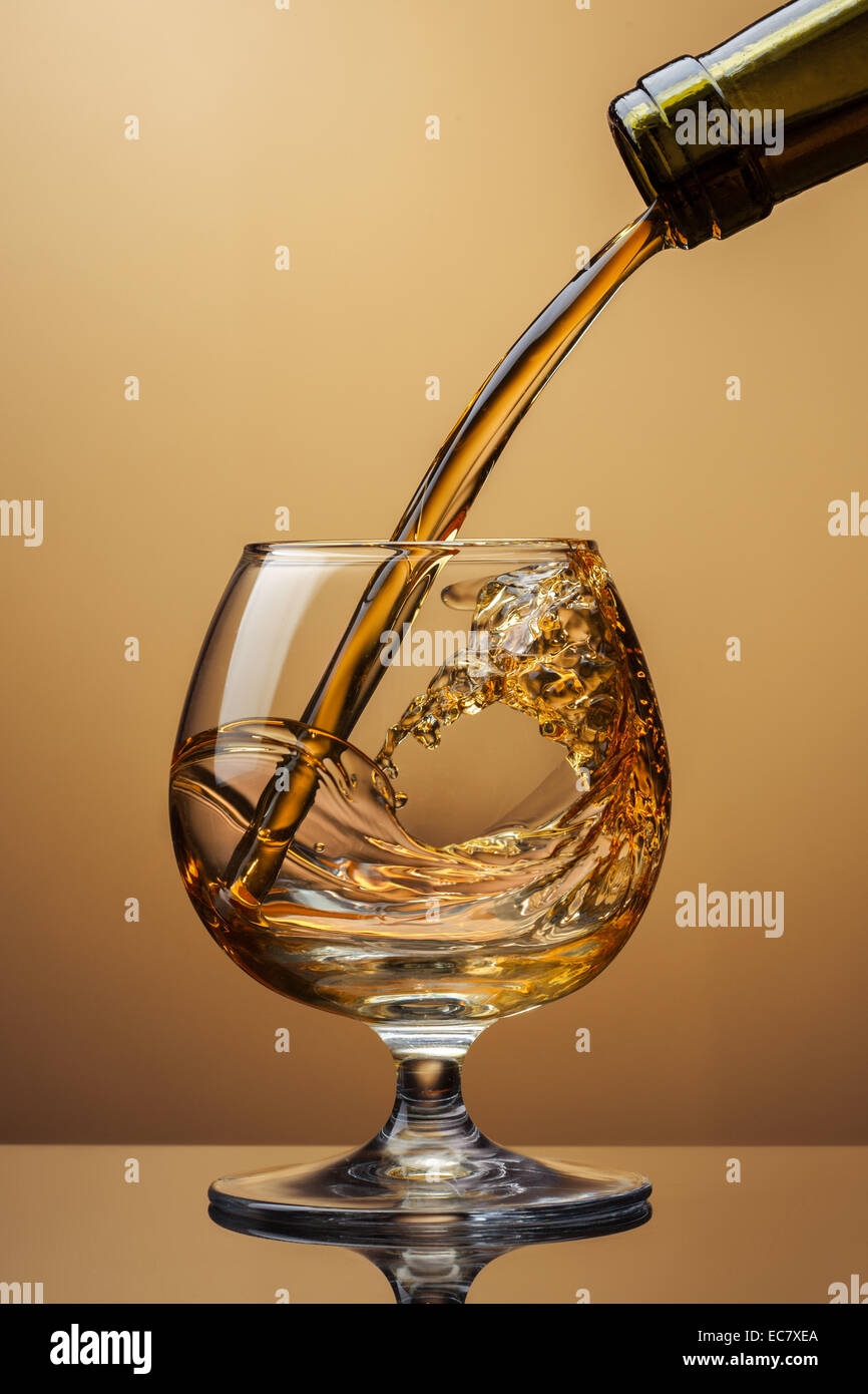Cognac pouring from bottle into glass with splash on brown background ...