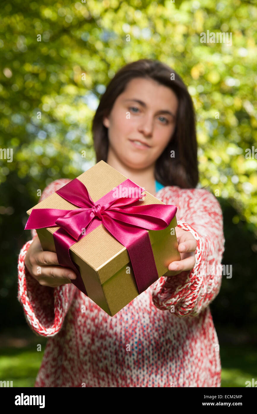 young woman offering golden red gift box over green tree background ...