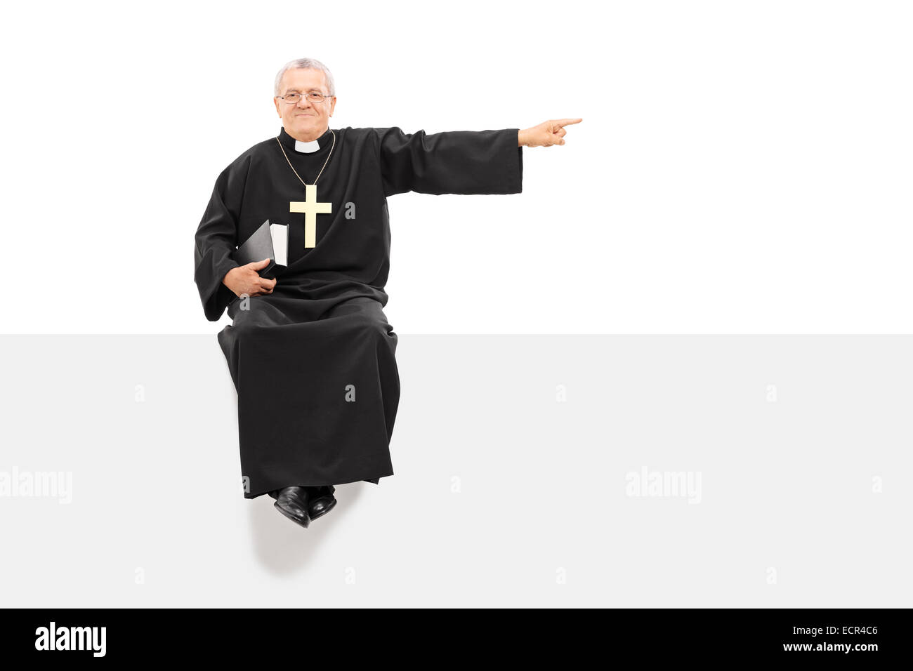 Mature priest pointing with his hand seated on panel isolated on white ...