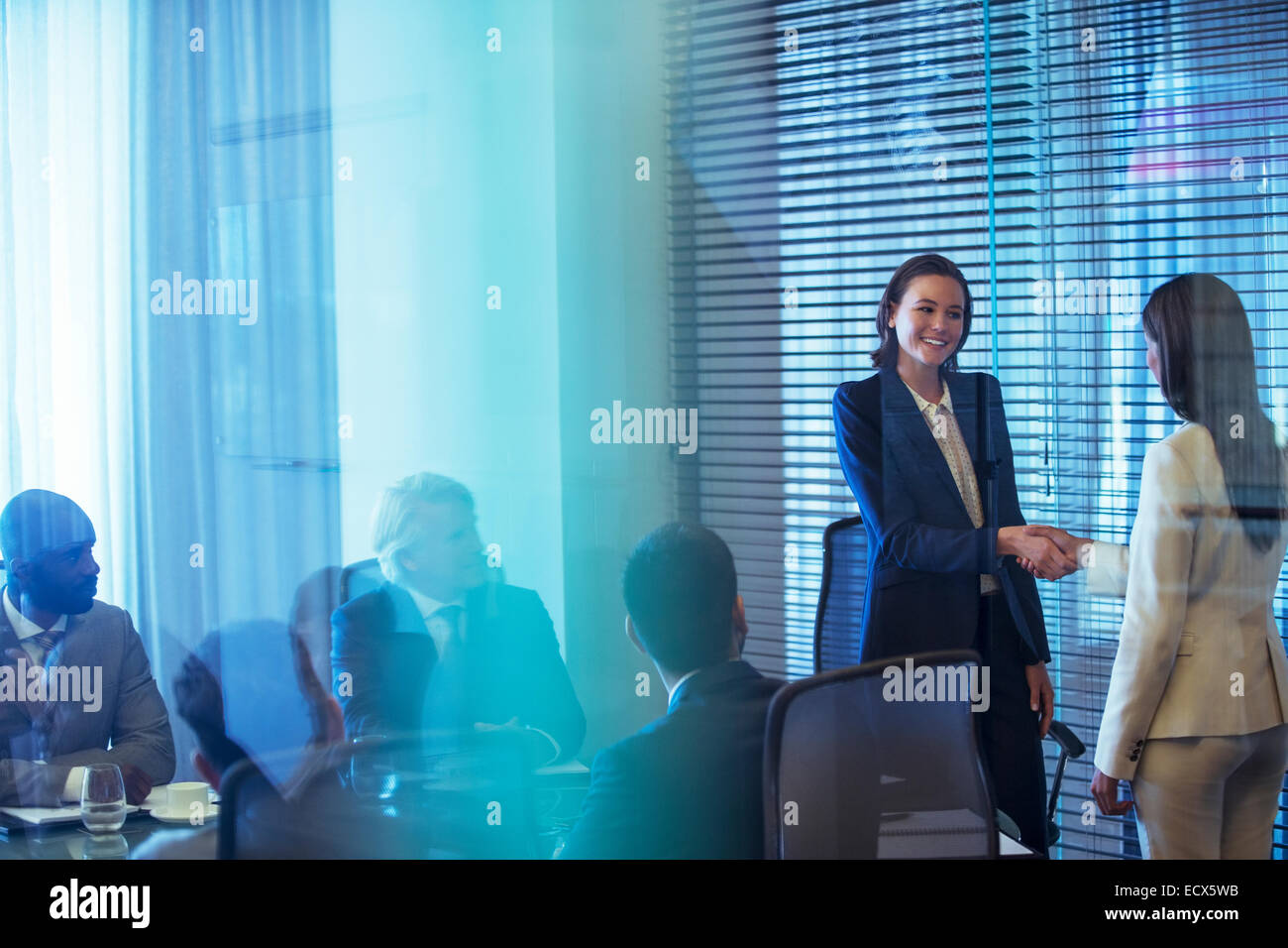 Two businesswomen shaking hands in conference room, colleagues watching ...