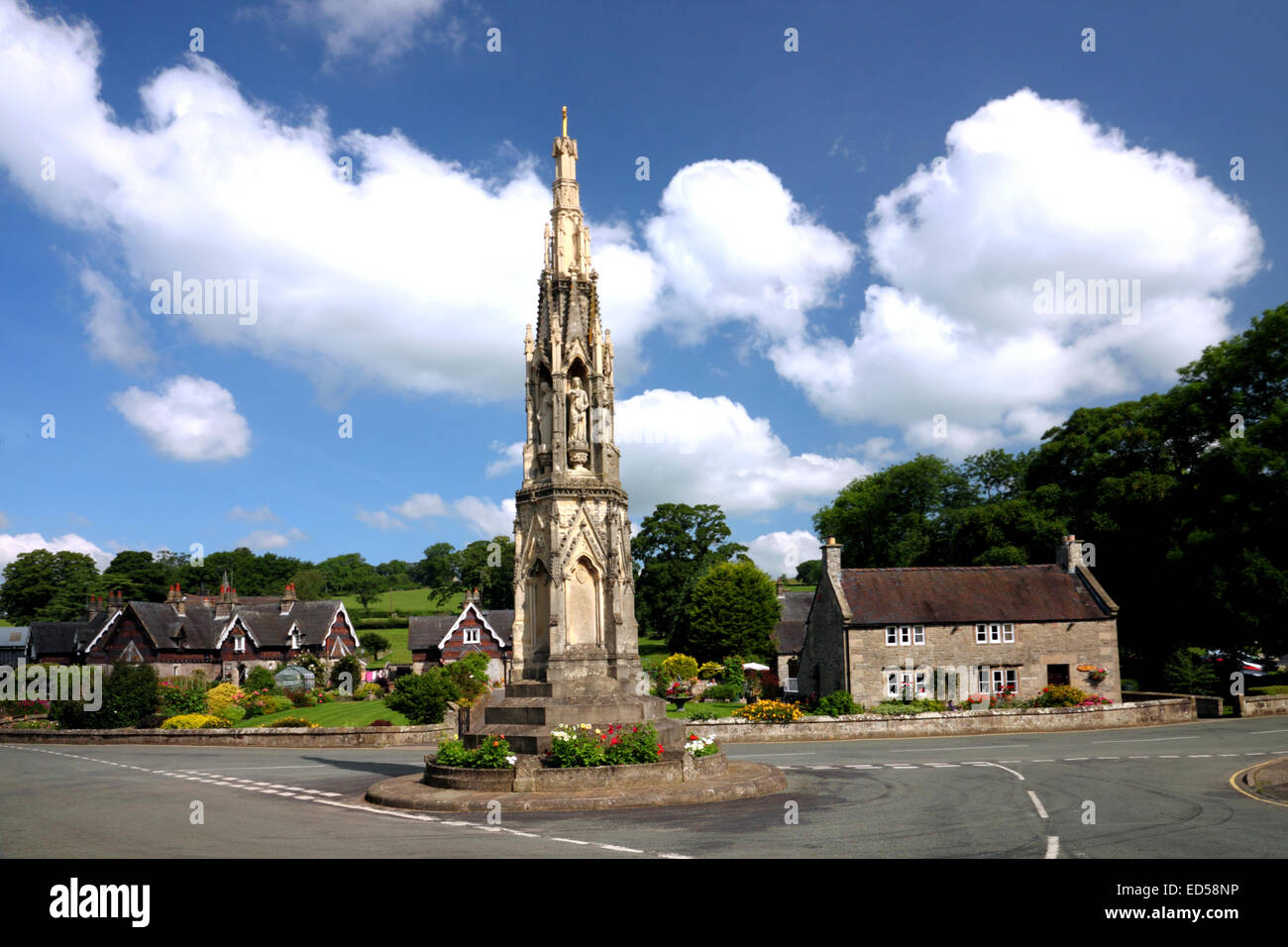 A tall Gothic monument on a roundabout with stone clad cottages behind ...