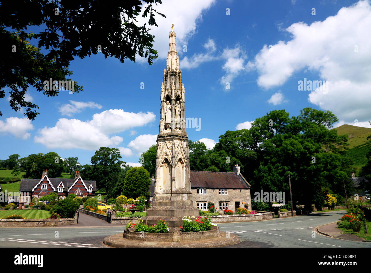 A tall Gothic monument on a roundabout with stone clad cottages behind ...