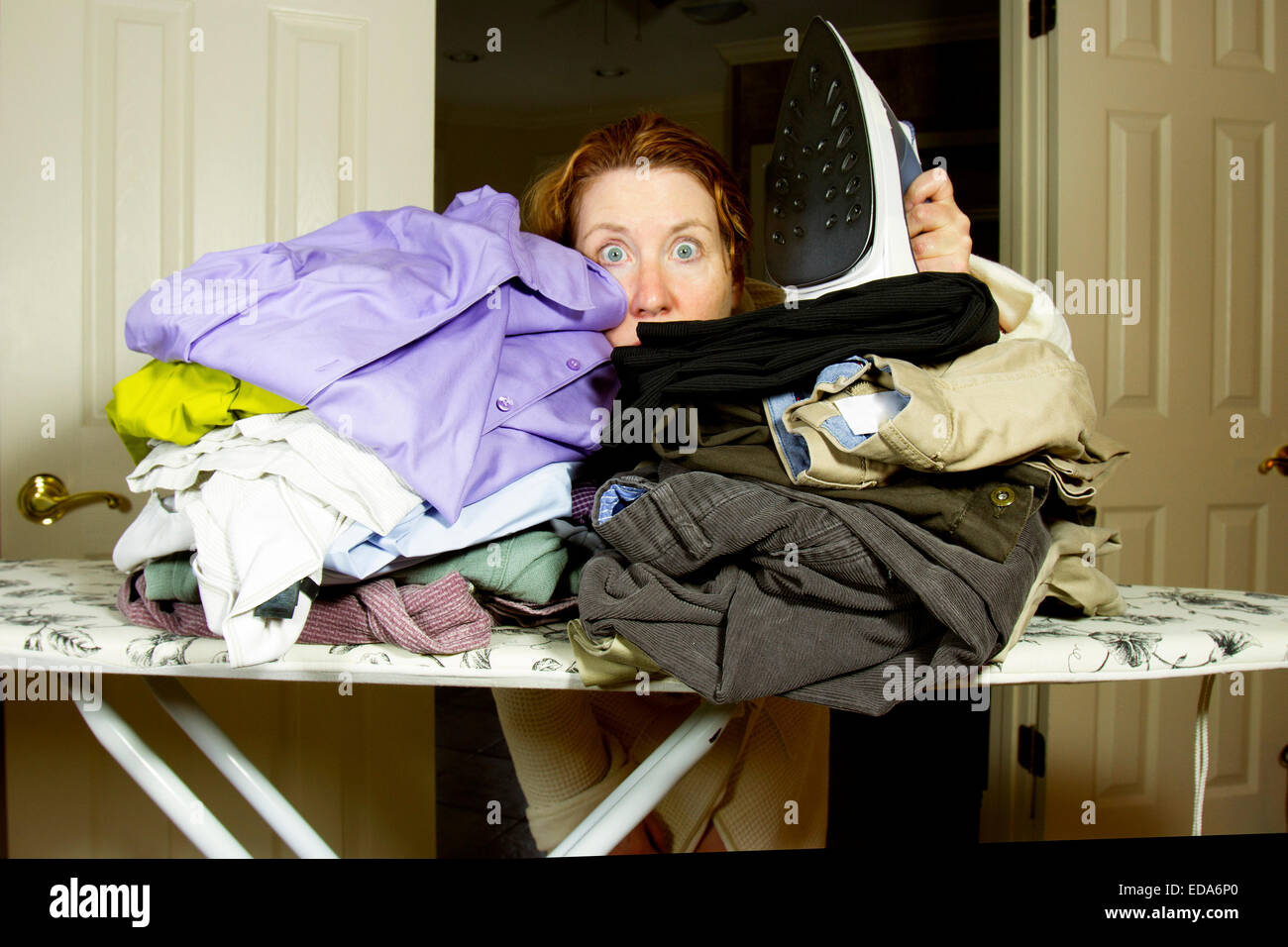 Woman peering through a pile of unironed clothes on an ironing board ...