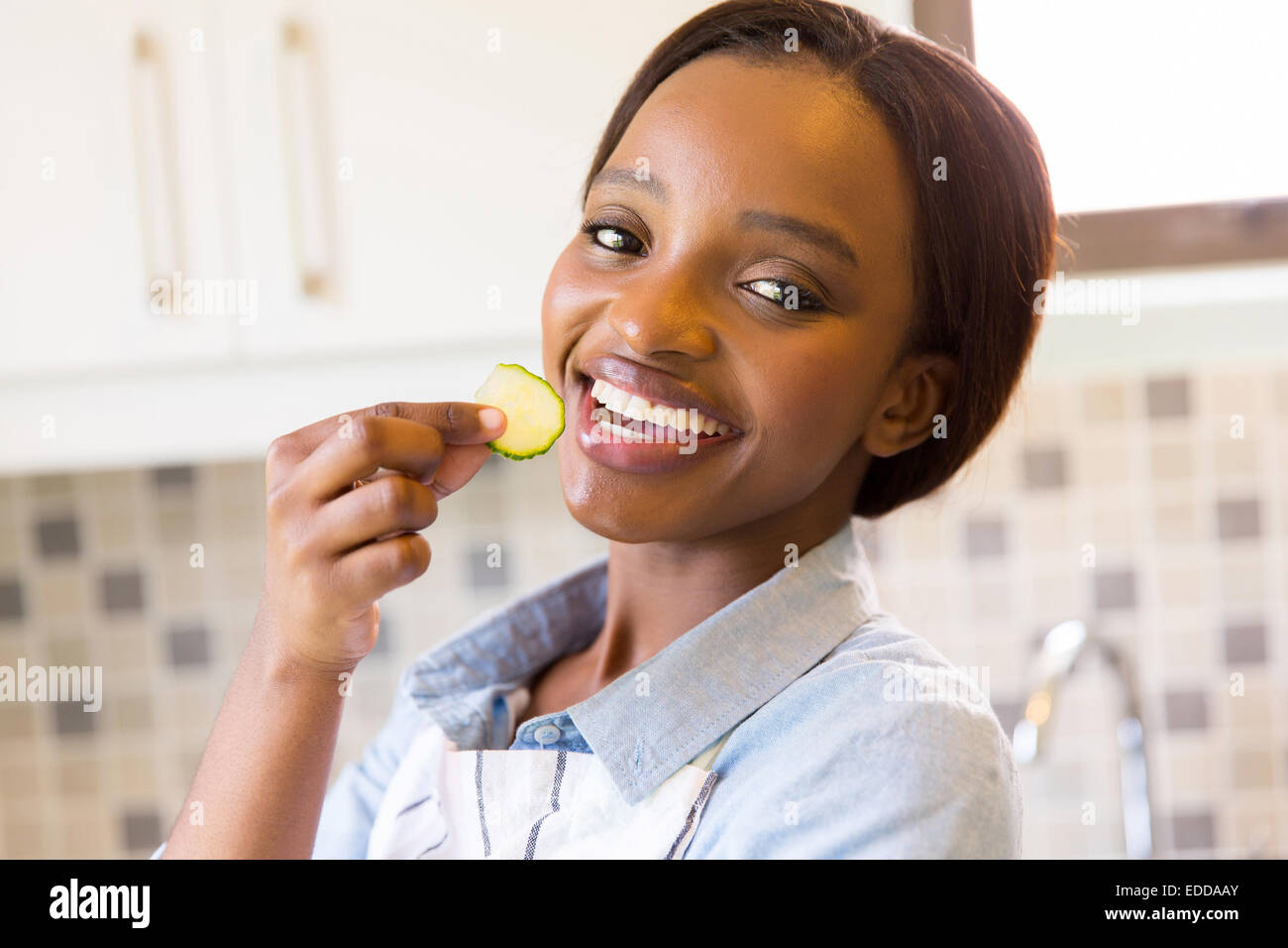 beautiful African woman eating a slice of cucumber in kitchen Stock ...