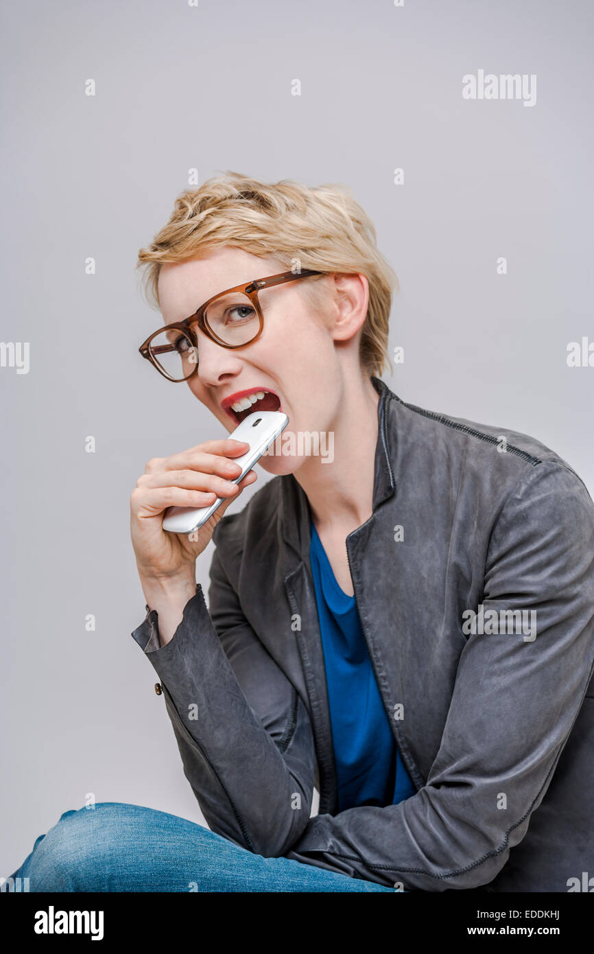 Portrait of blond woman biting her smartphone in front of grey ...