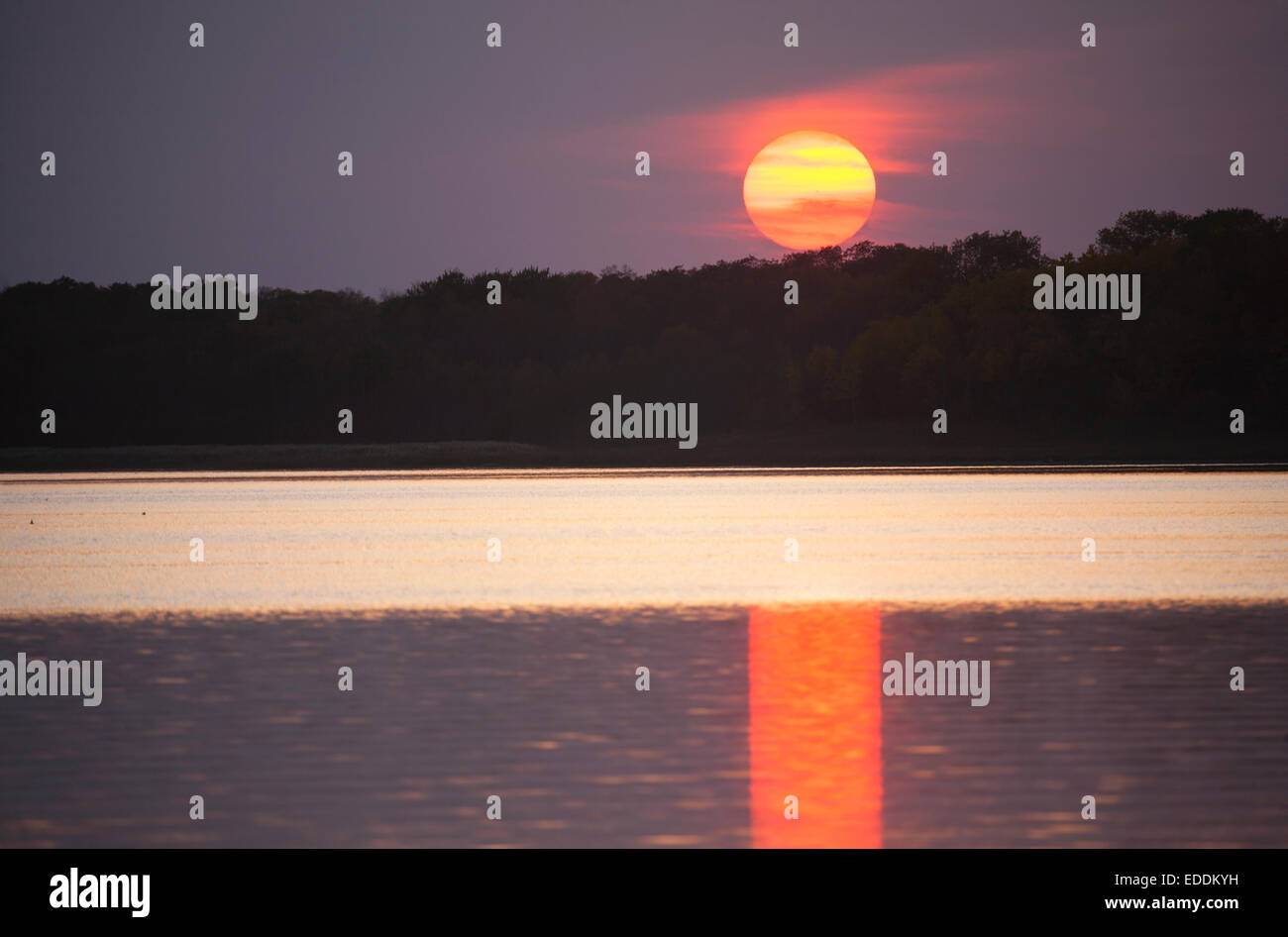 An orange sun sinking below the horizon, reflected in the water of a ...