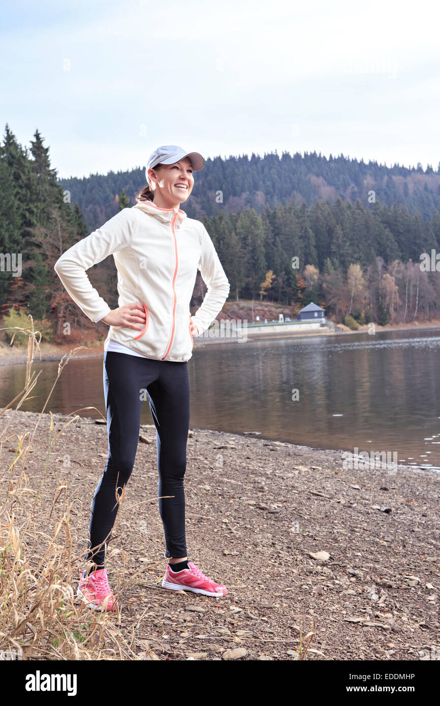 Smiling female jogger with hands in her hips standing at lakeshore ...