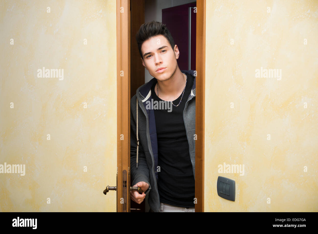 Handsome young man opening door to enter into a room, looking at camera ...
