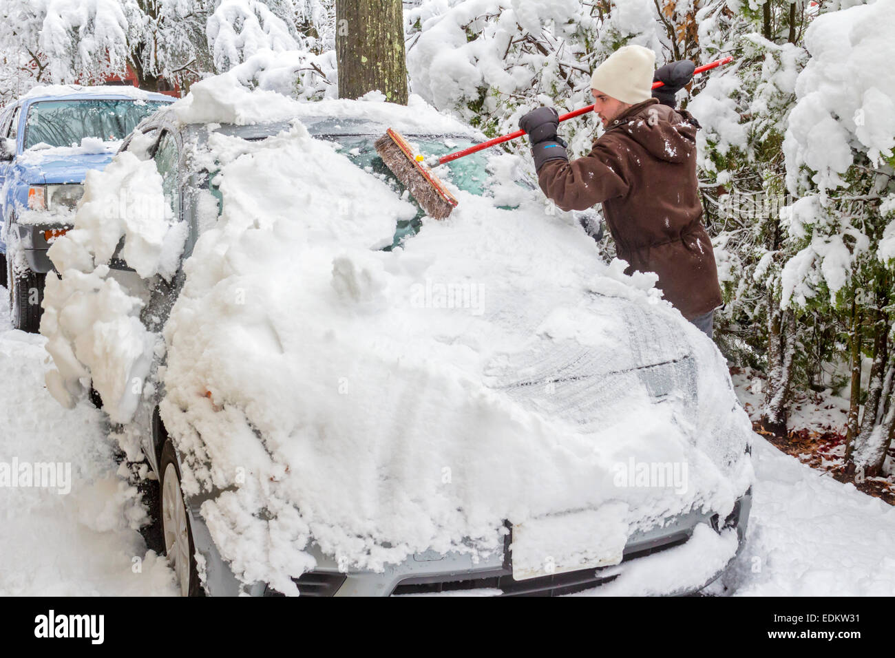 Young man dressed in a winter coat, hat and gloves cleans snow from car ...