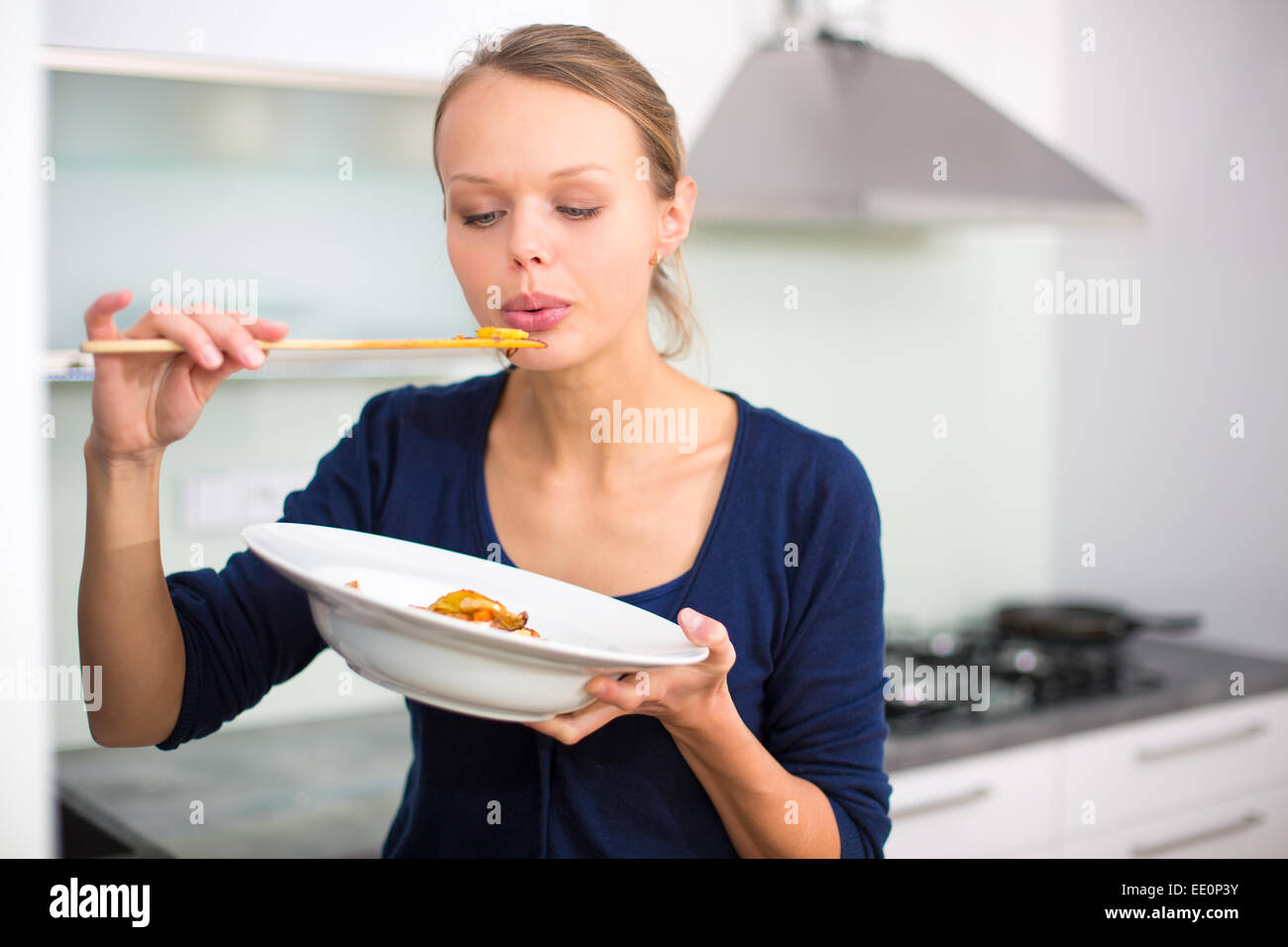 Pretty, young woman cooking a diner in a modern kitchen, tasting the ...
