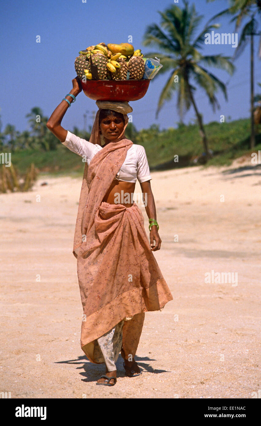 Fruit seller with fruit in container on her head, palm tree behind ...