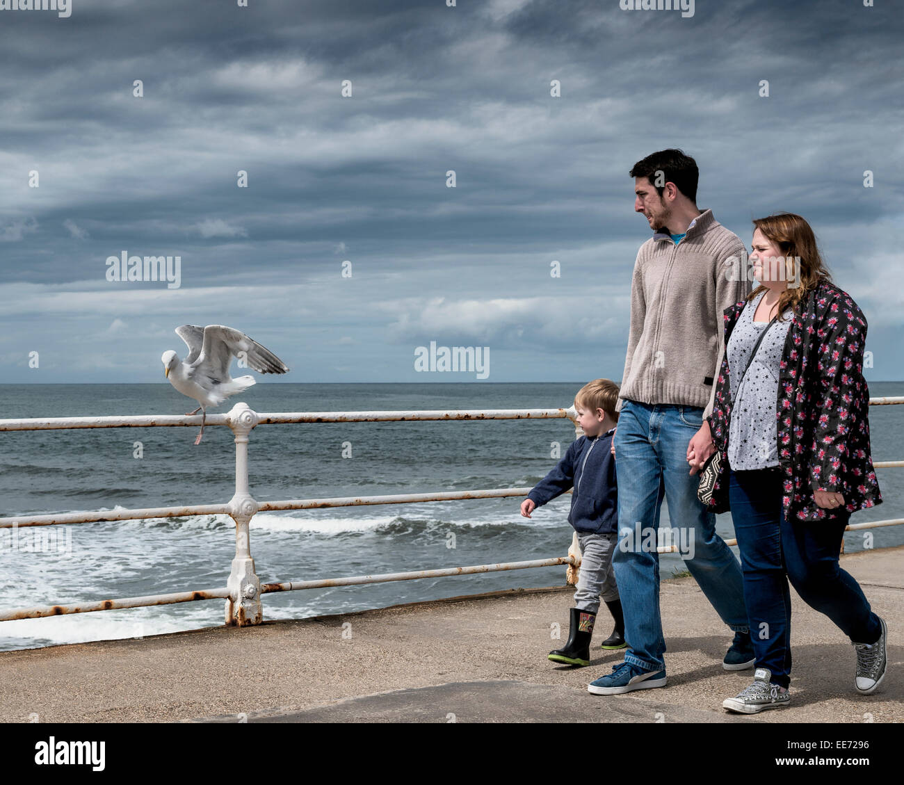 A young family walk along a British seaside pier, holding hands on a ...