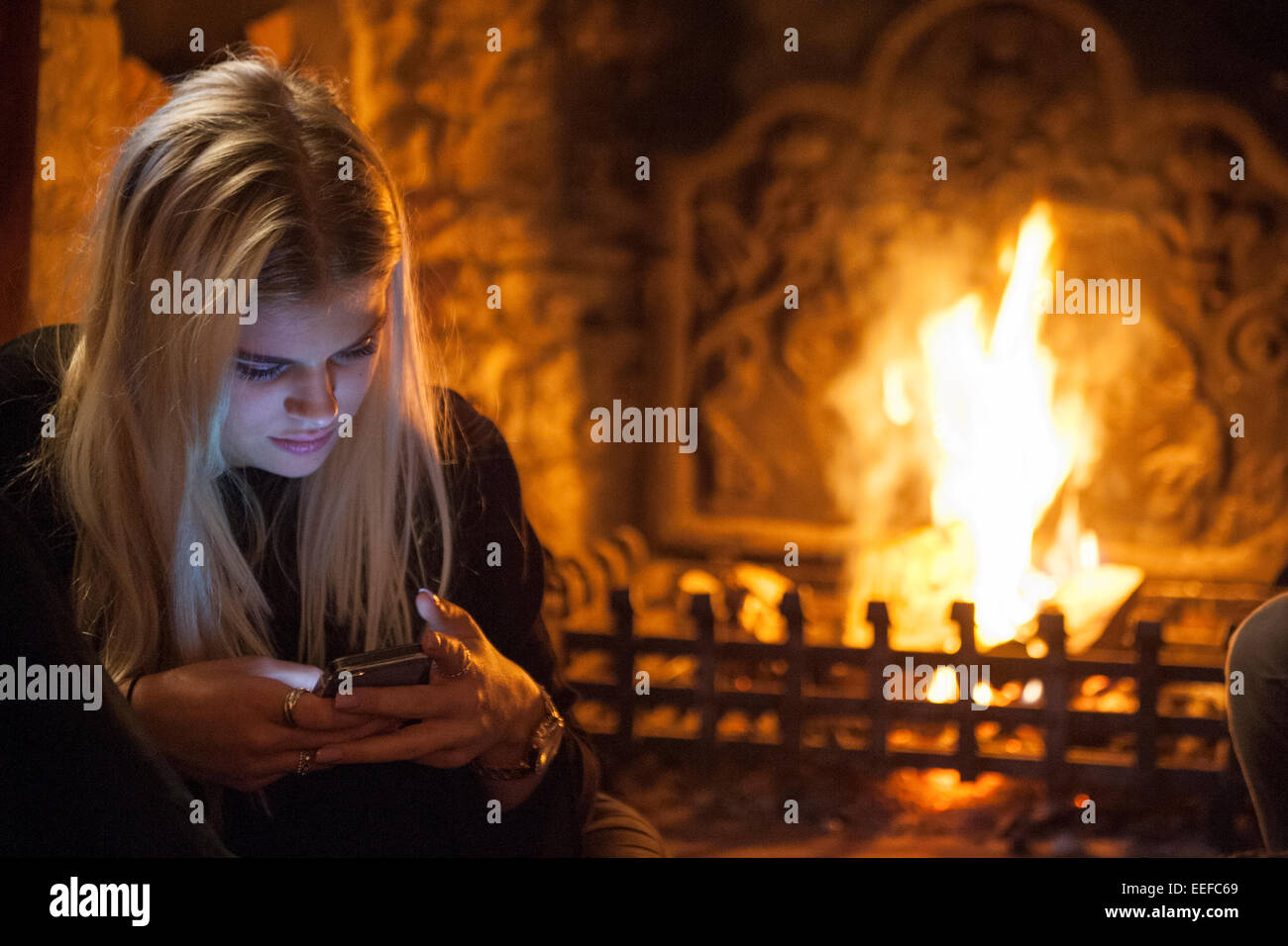 A girl checks her mobile phone whist sitting in front of a roaring fire ...