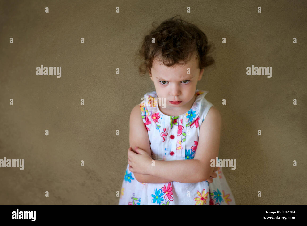 Grumpy girl (2-3) in floral patterned dress standing by wall with arms ...