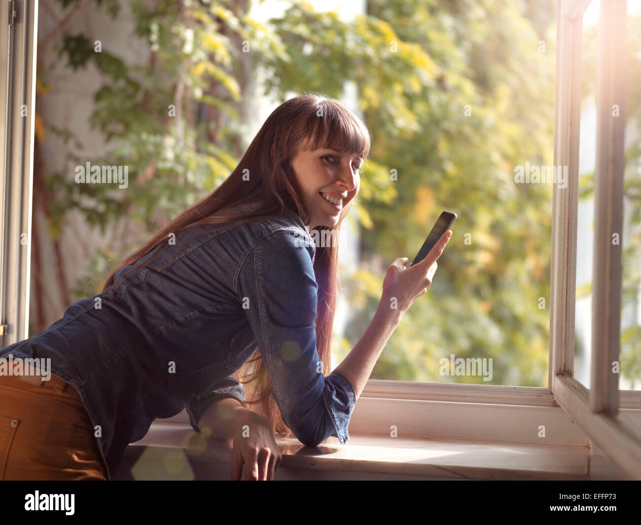 Smiling woman at open window holding cell phone Stock Photo - Alamy