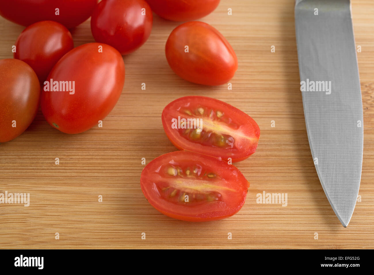 A single cut grape tomato next to a knife with more tomatoes in the ...