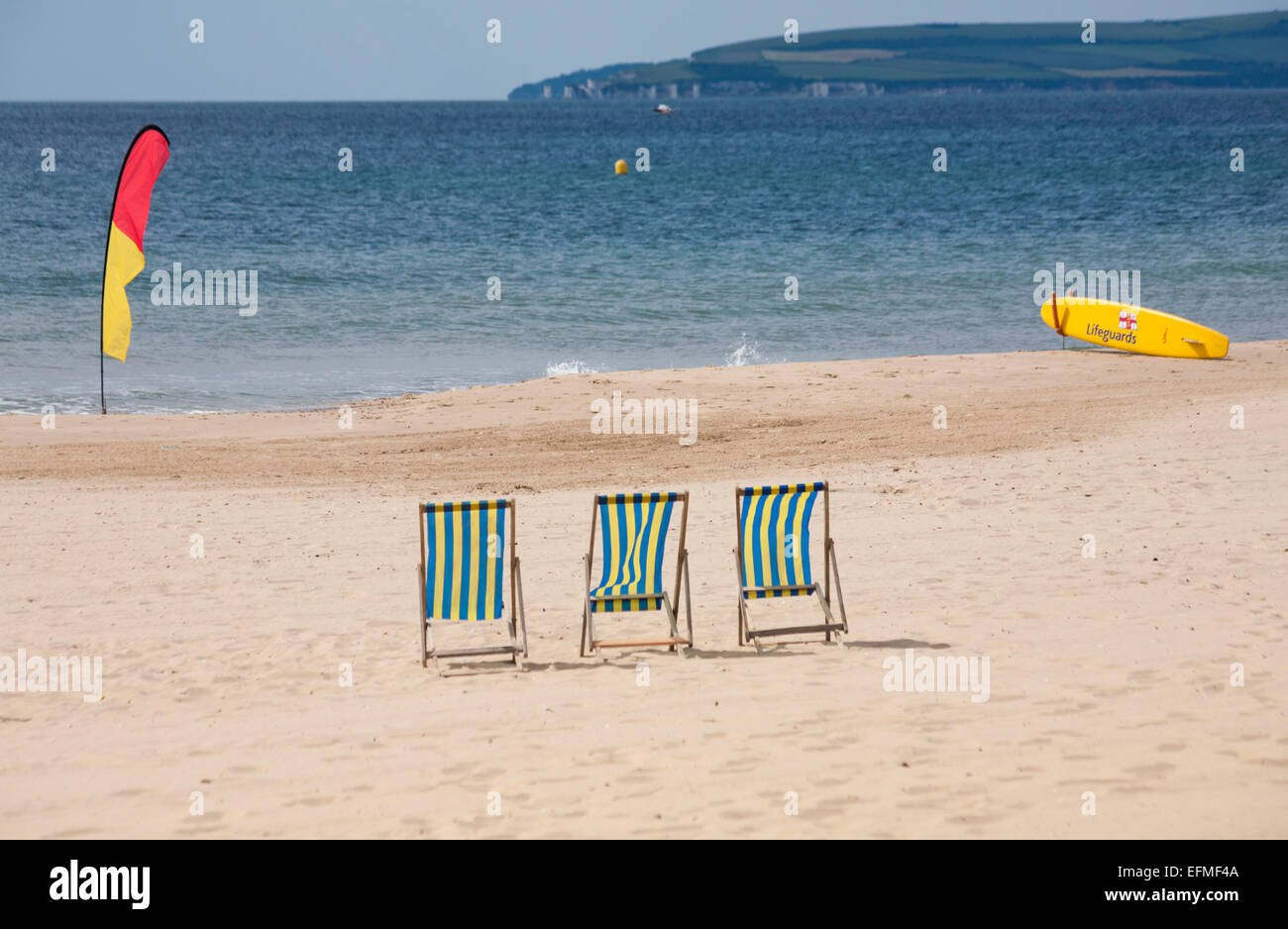 Three empty deckchairs between RNLI Lifeguards surf board and marker ...