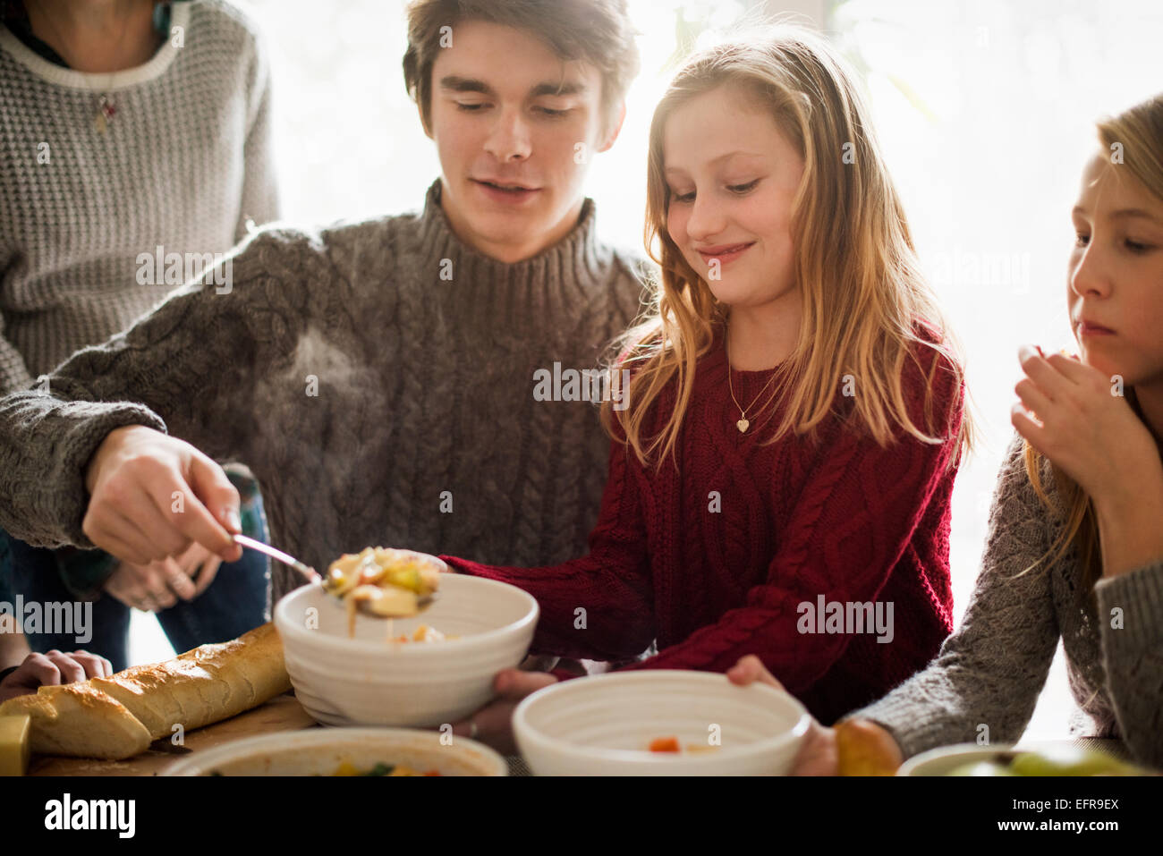Four people sitting and standing at a table, a man serving food into a ...
