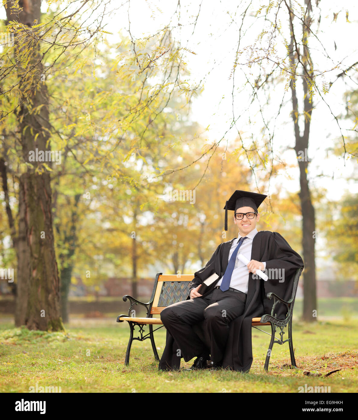 Graduate student holding diploma and a book seated on bench in park ...