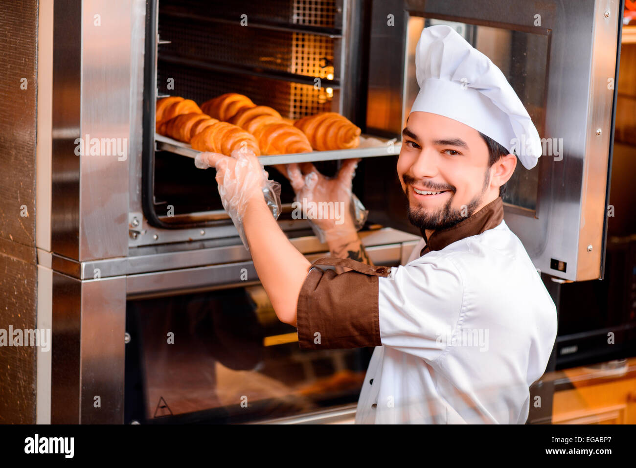 Confident baker at work. Cheerful young baker in apron putting g a tray ...