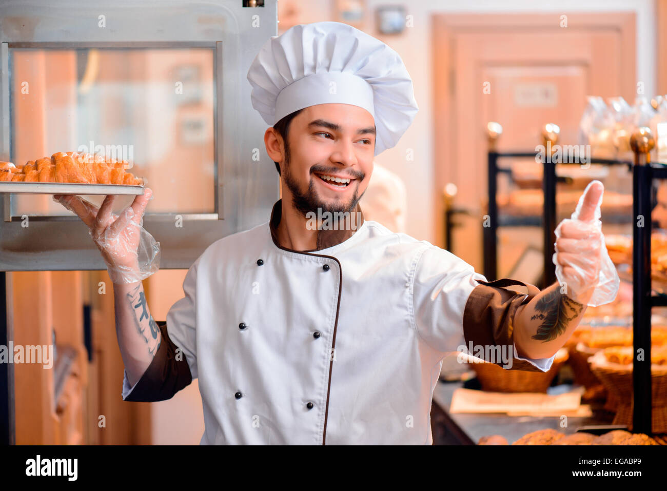 Confident baker at work. Cheerful young baker in apron putting g a tray ...