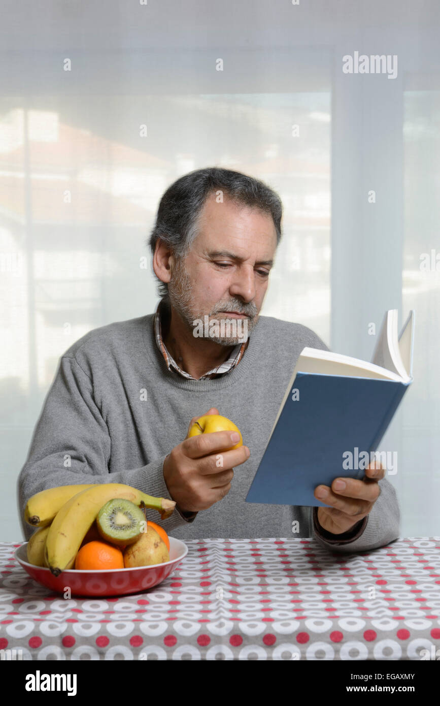 Middle aged man reading a book while eating fruit for breakfast in the ...