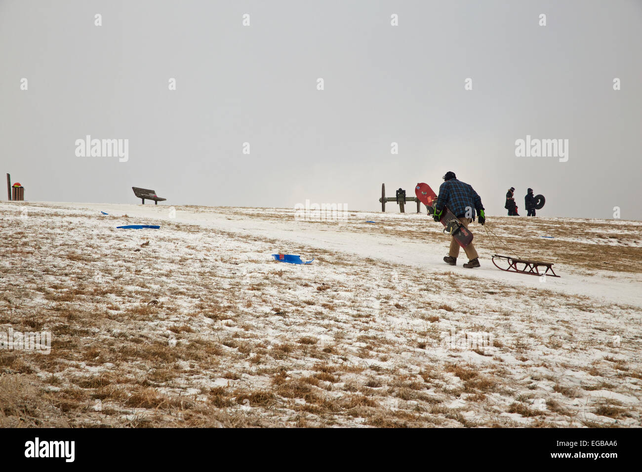 Man pulling sled and carrying a snowboard to the top of a snowy hill ...