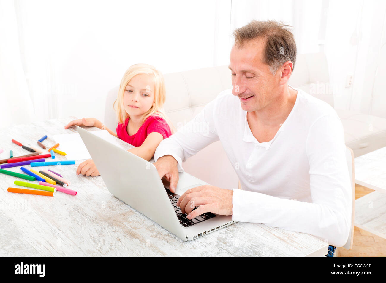 A father working on a laptop computer while his daughter is drawing ...