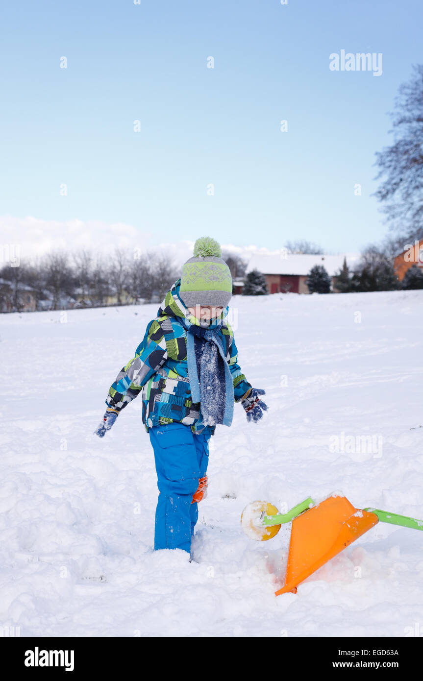 Young child boy plays at backyard collecting snow by wheelbarrow toy ...