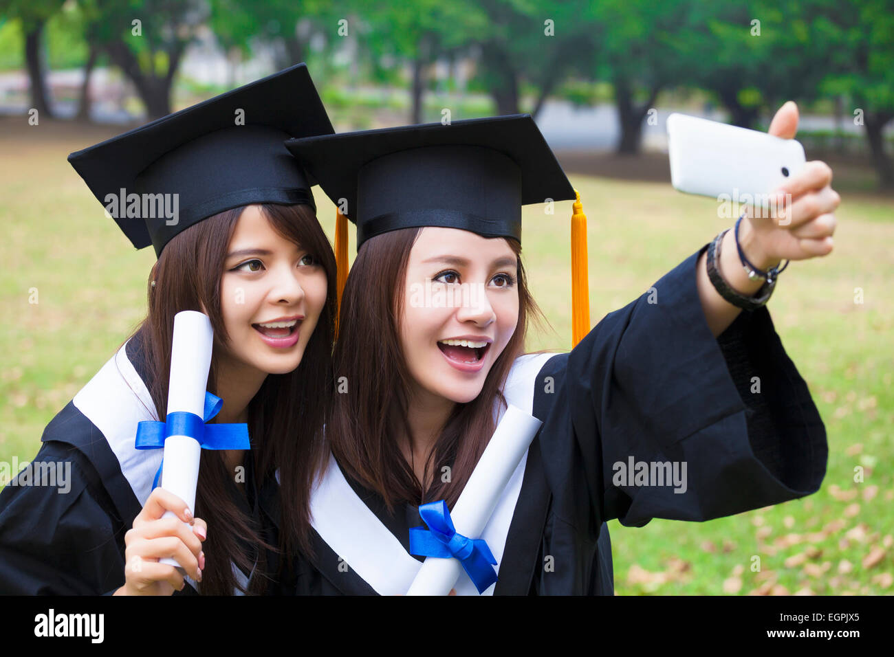 Two happy women in graduation gowns taking picture with cell phone ...