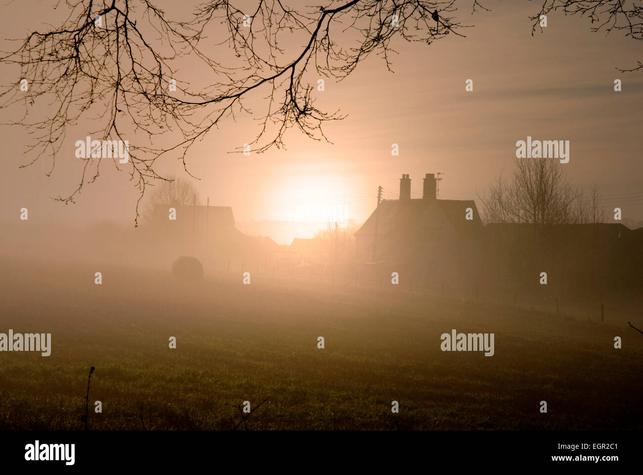 Dawn - rising sun shrouded by mist - farm buildings in dark silhouette ...