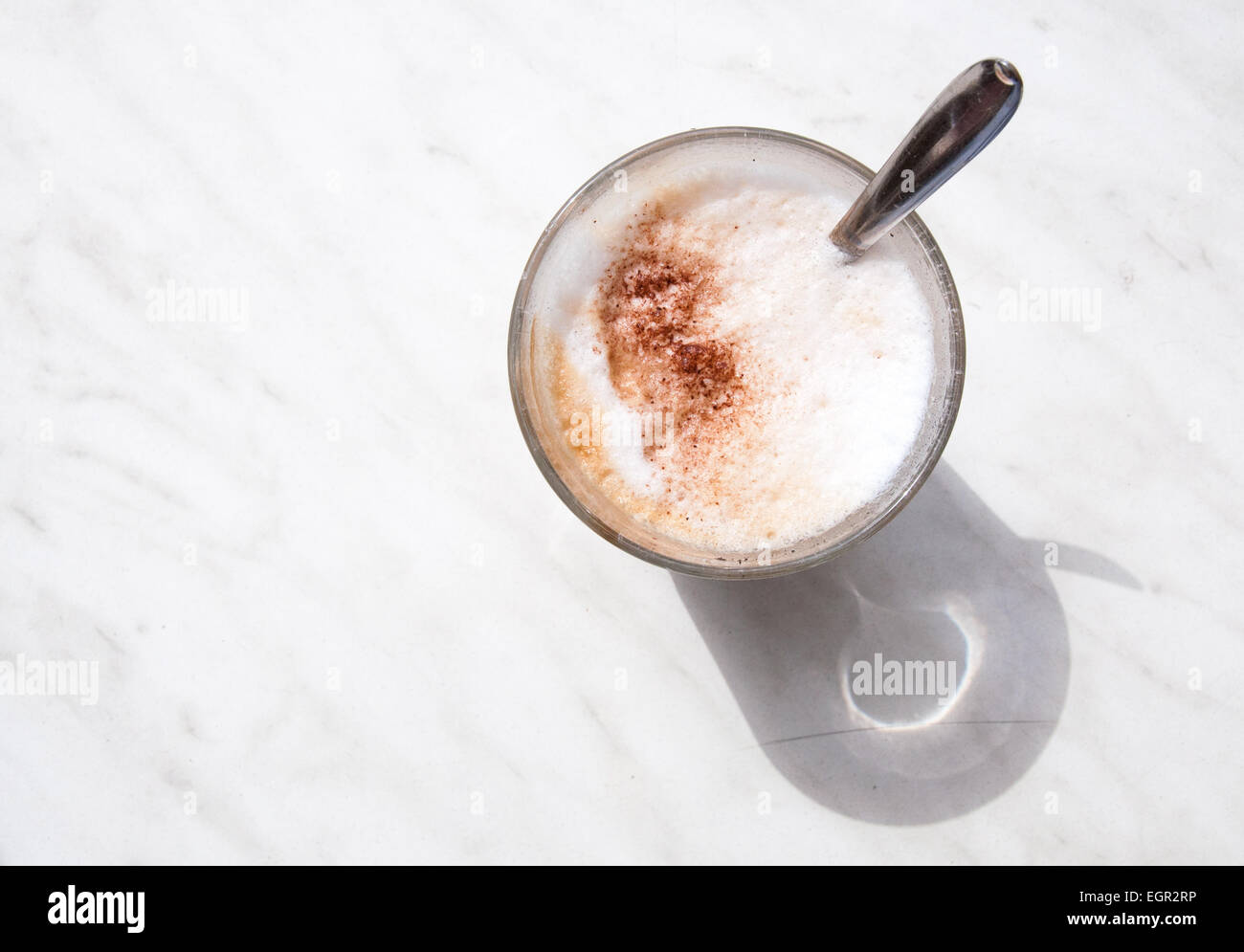 Foamy cappuchino in a coffee glass with spoon on white marble surface ...