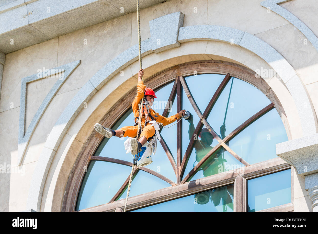 Window cleaner wearing orange overalls and red helmet, suspended from ...