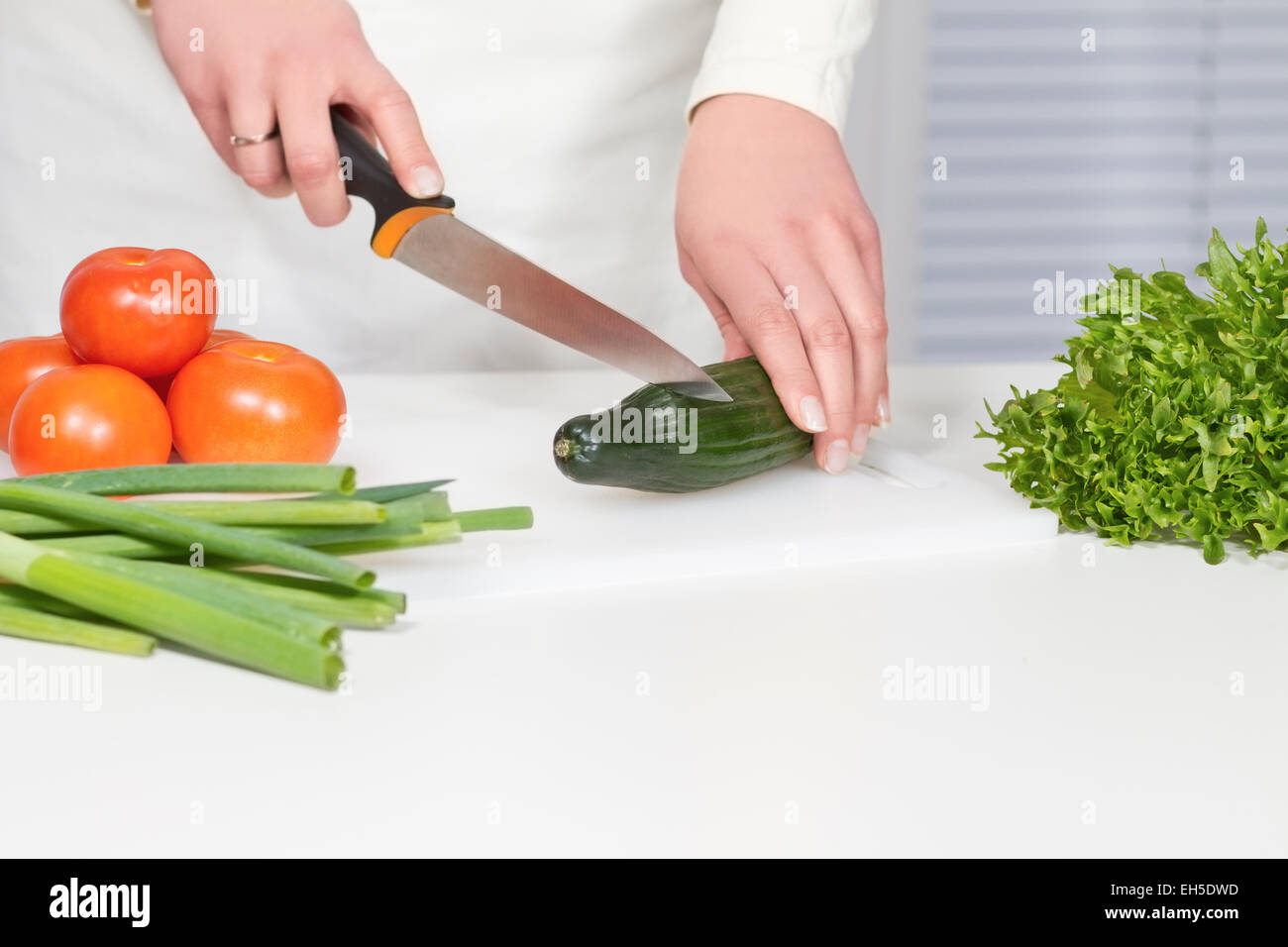 Woman in a white kitchen chopping a cucumber to a salad with a knife ...