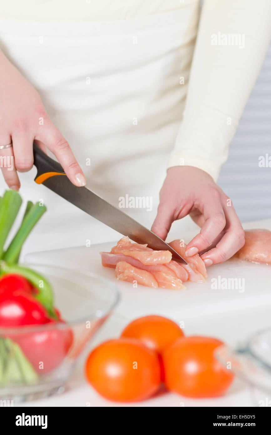 Young woman in a white kitchen chopping chicken meat fillet. Vegetable ...