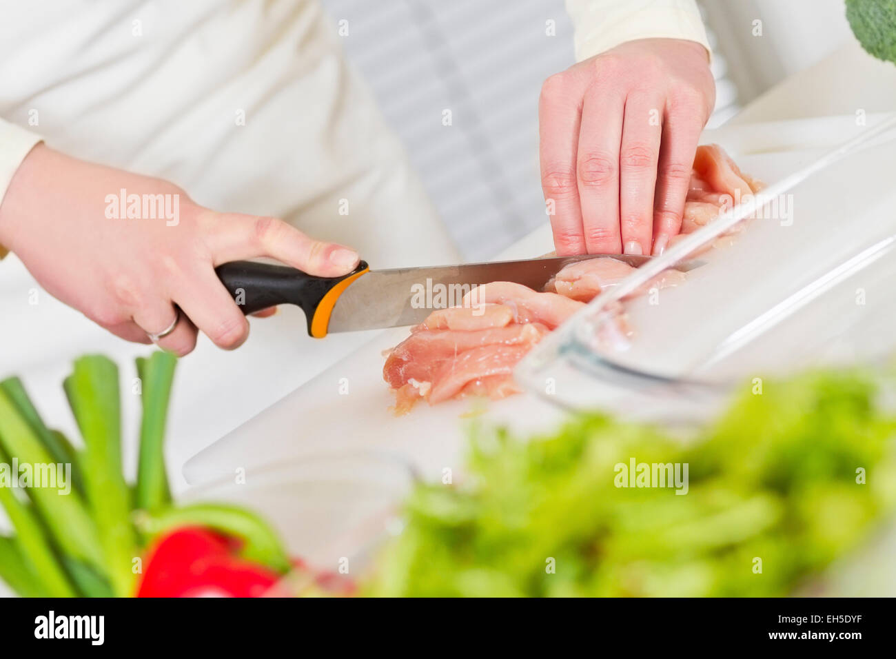 Young woman in a white kitchen chopping chicken meat fillet. Vegetable ...